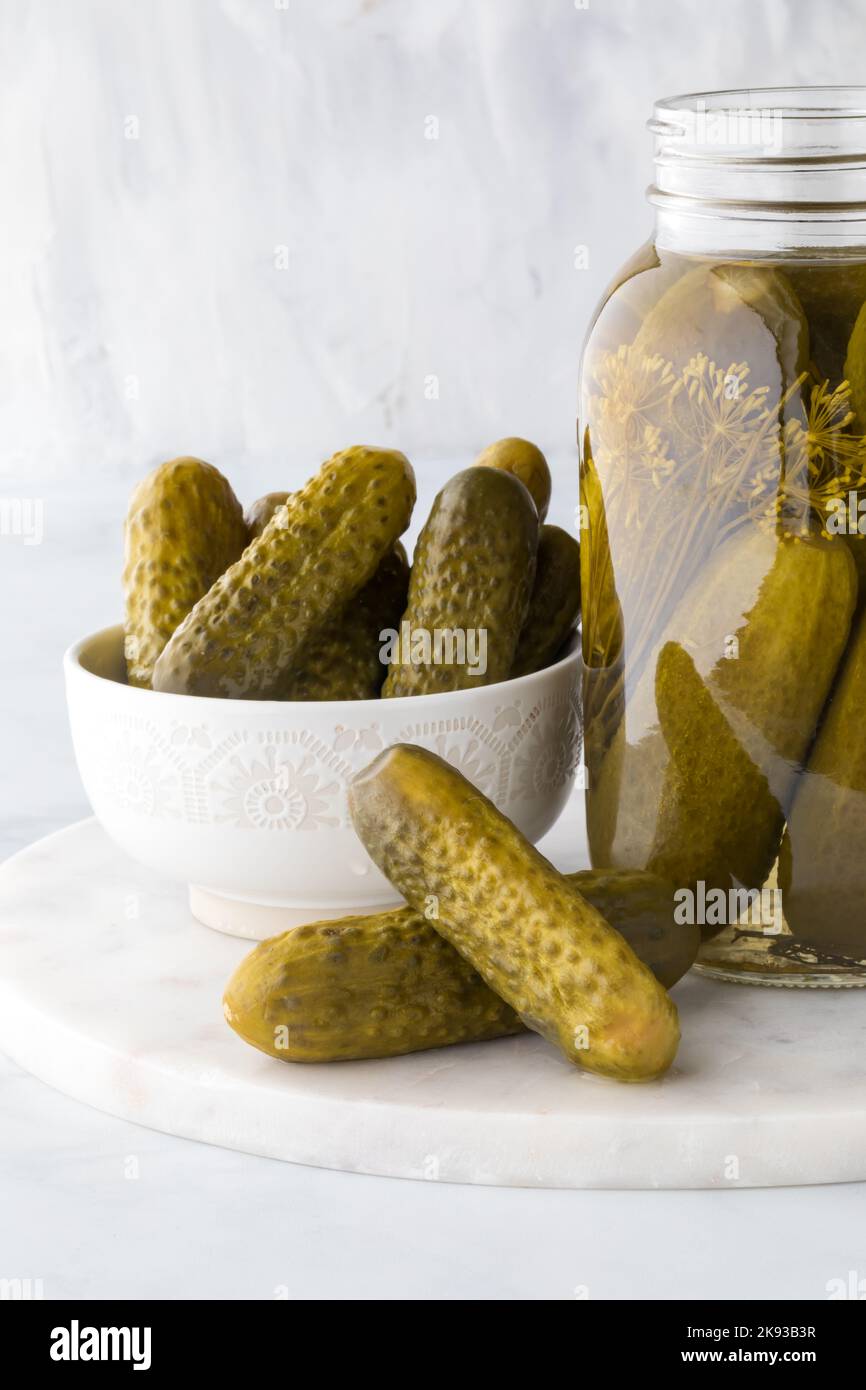 A bowl and jar of dill pickles with two pickles in front on a marble ...
