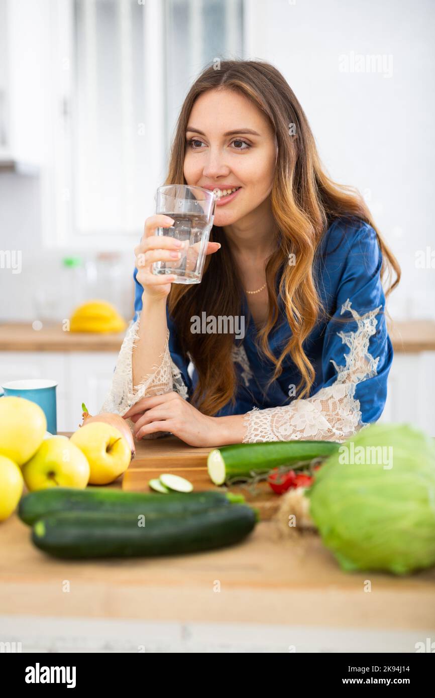 Smiling young woman in blue dressing gown drinking water in kitchen ...