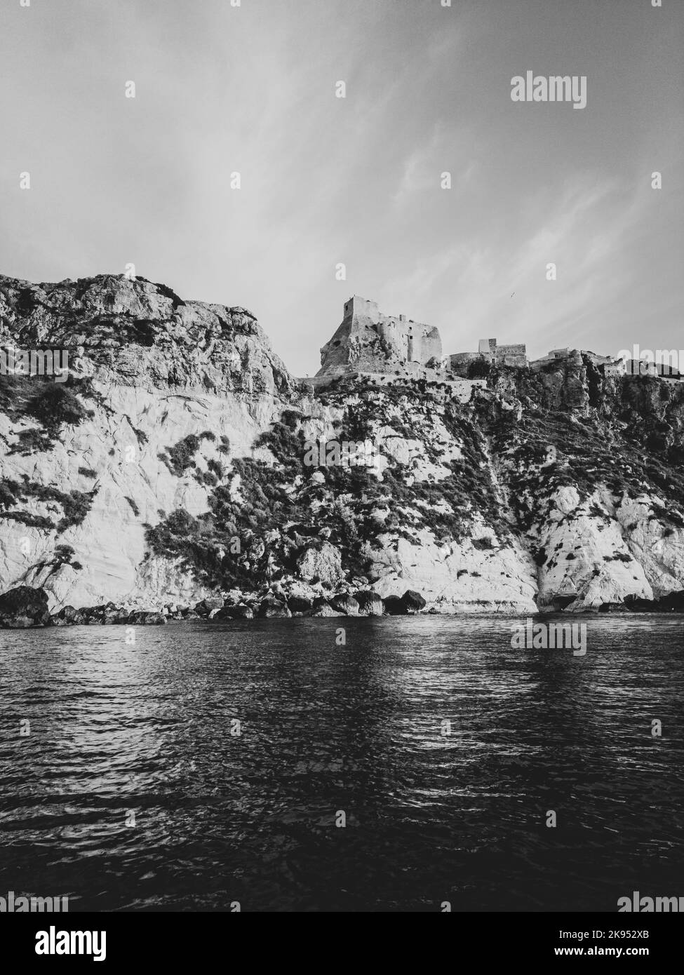 A vertical grayscale shot of castle ruins on a cliff over water at the ...