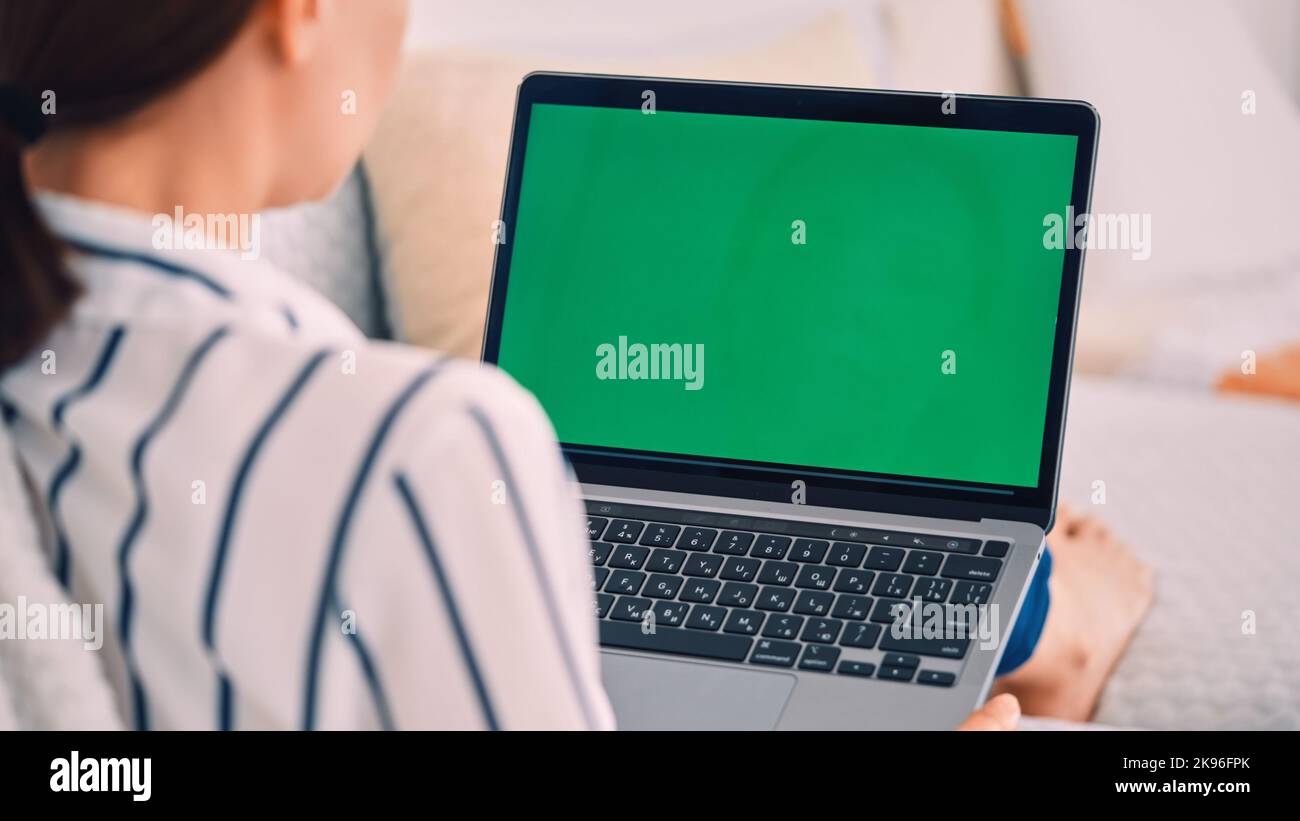A woman sitting on the couch and using a laptop with a green screen ...