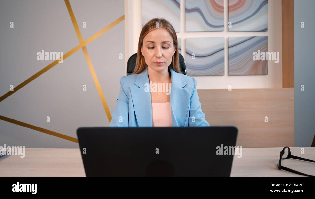 A Caucasian woman in a formal outfit working at her desk using a laptop ...
