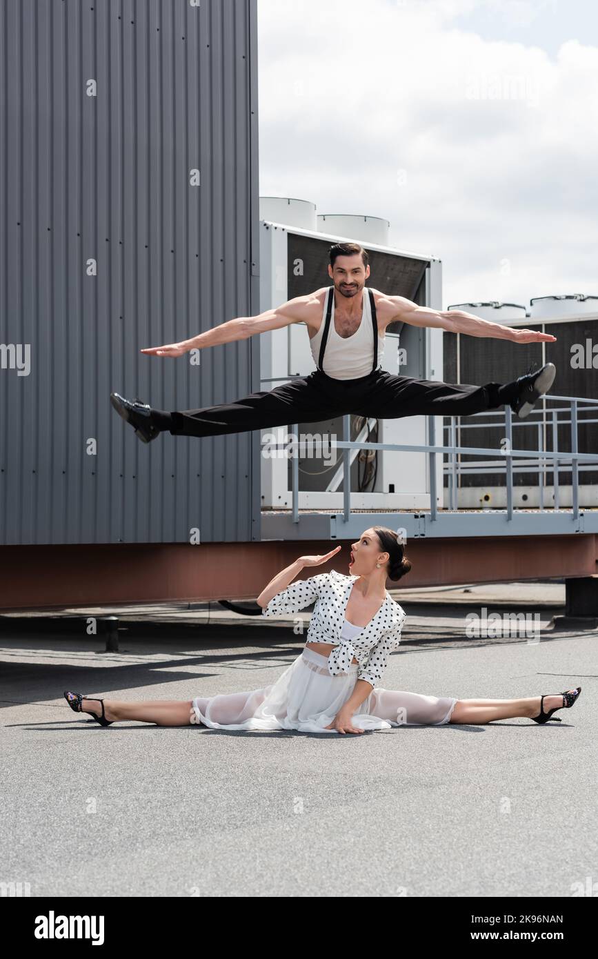 Smiling dancer doing split while jumping above shocked partner on roof ...