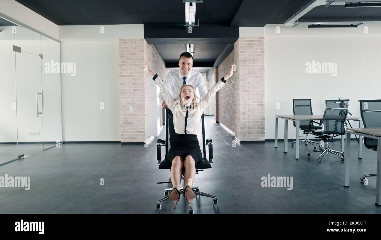 A male employee pushing his female coworker on a roller chair, having ...