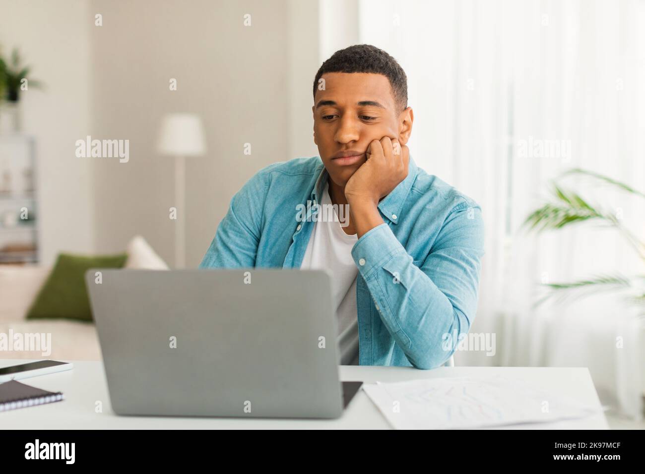 Sad tired bored young black guy in casual looks at laptop in light ...