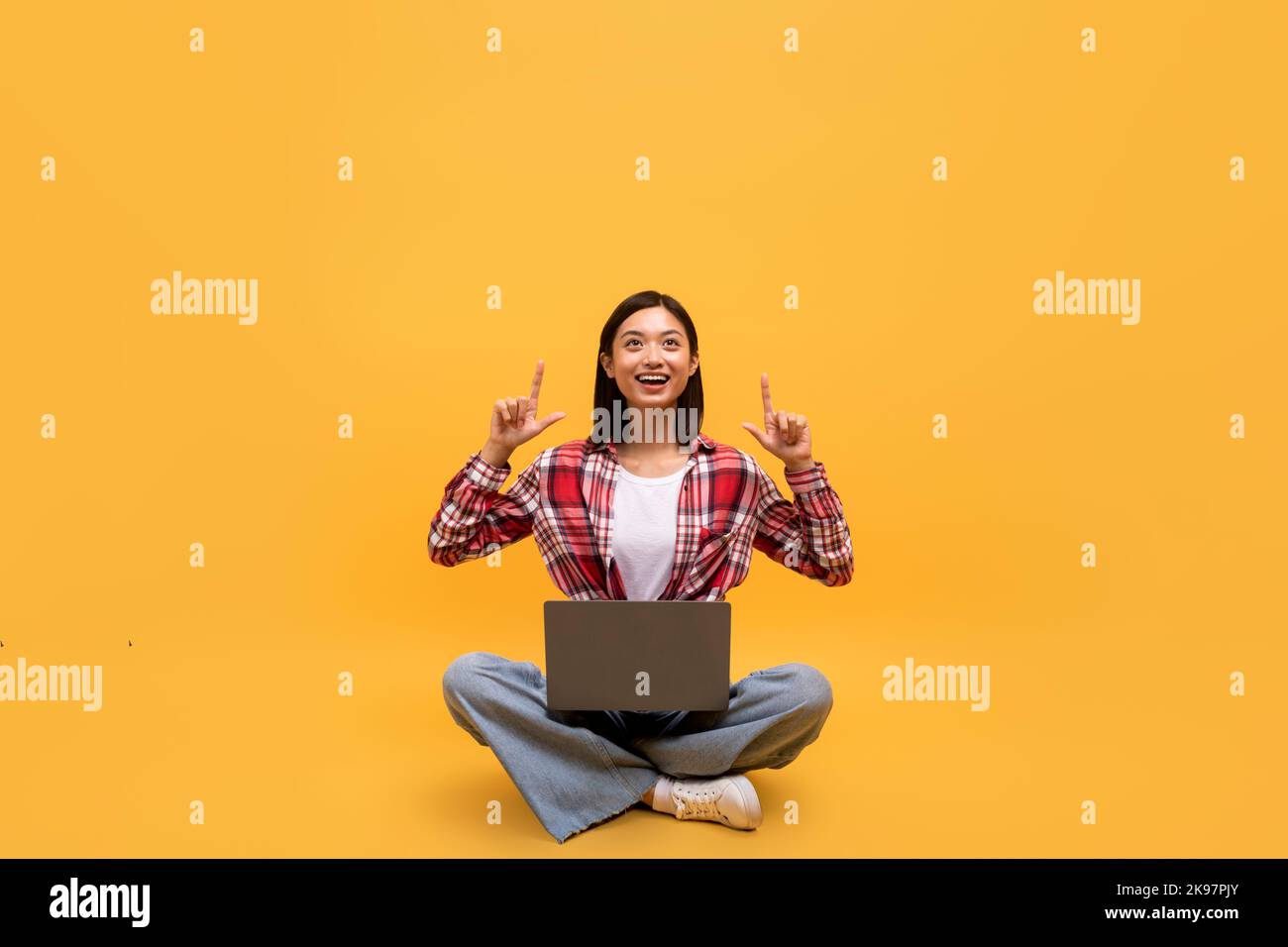 Happy asian lady sitting cross legged with laptop, pointing up at empty ...