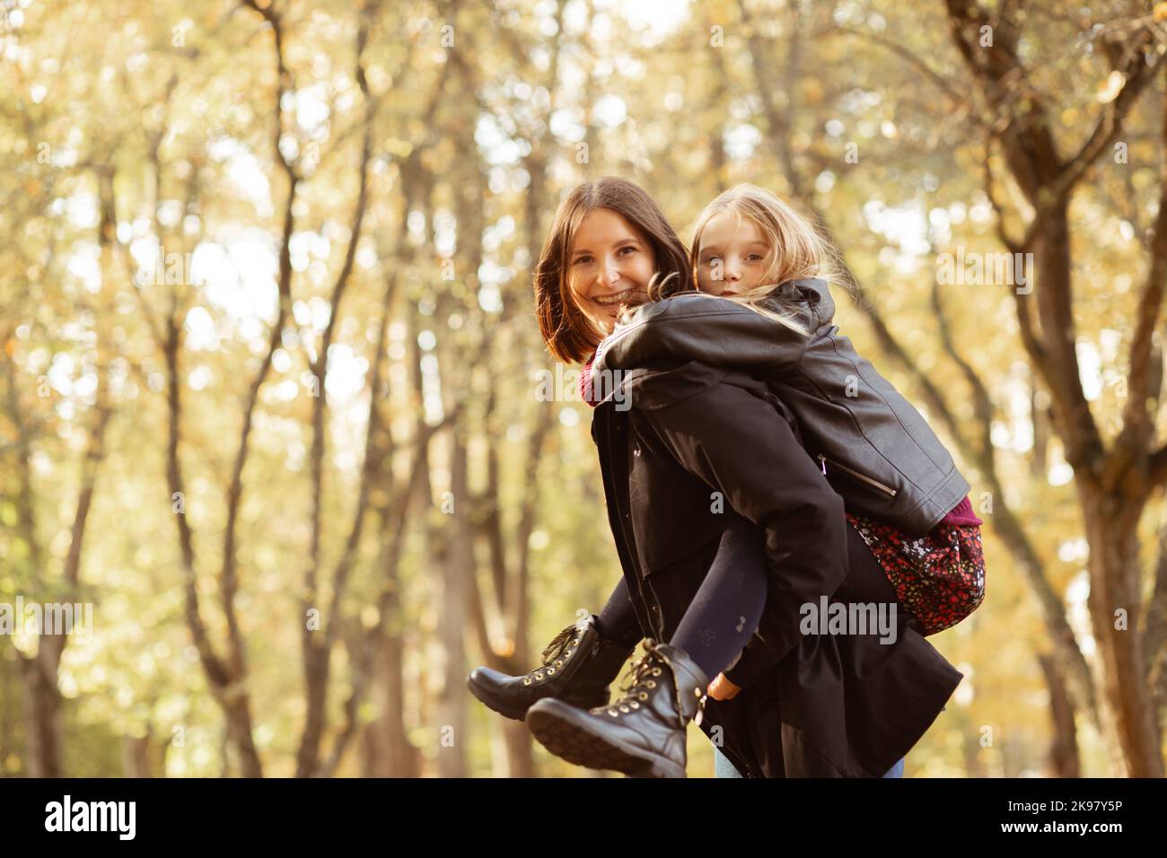 Joyful young woman ride on back little girl side view. Happy mother and ...