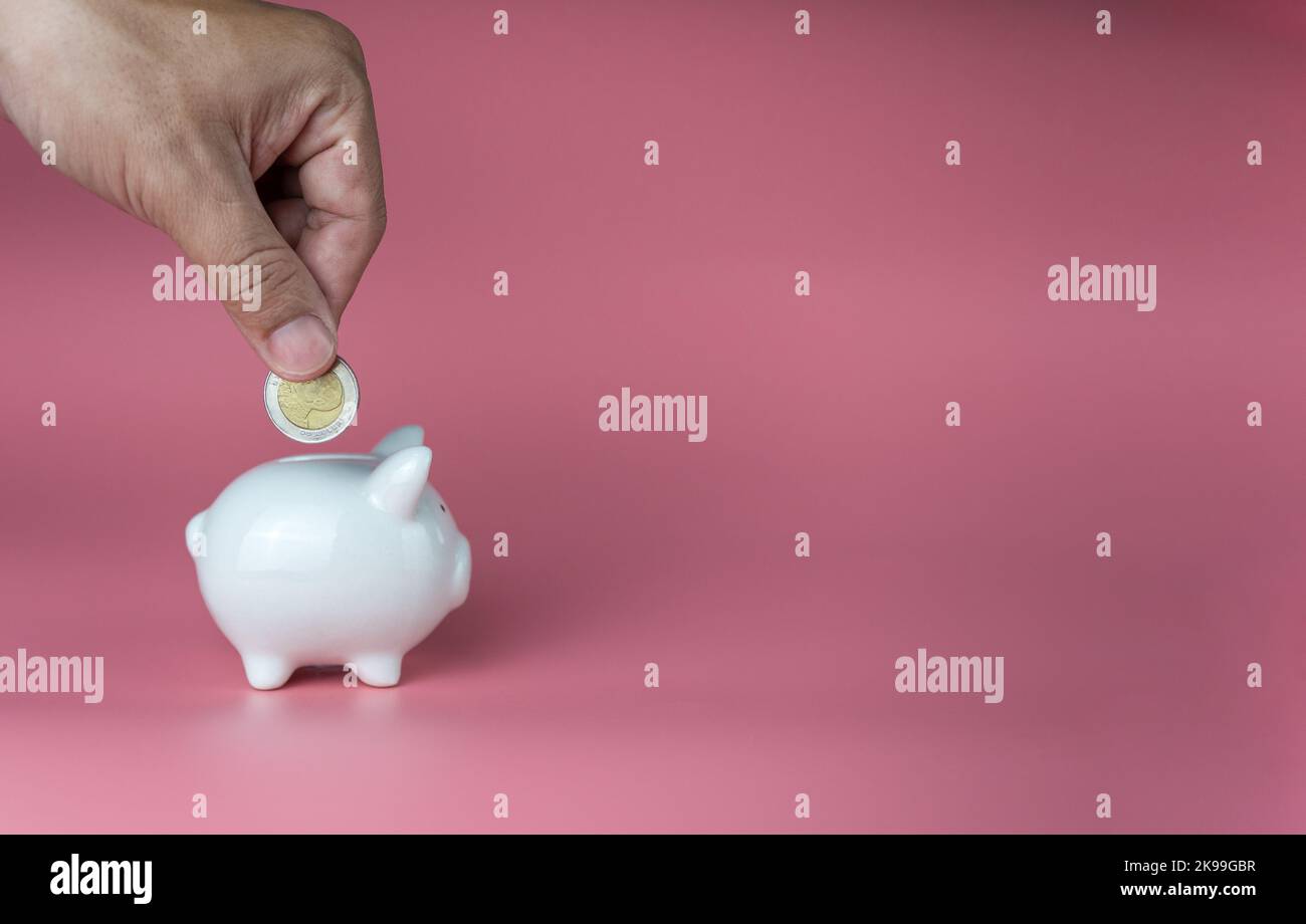 A human hand put a coin in a piggy bank on a pink background. Savings ...