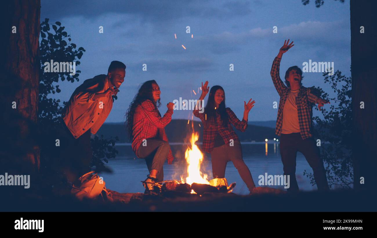 joyful friends multiethnic group are dancing around fire in forest ...