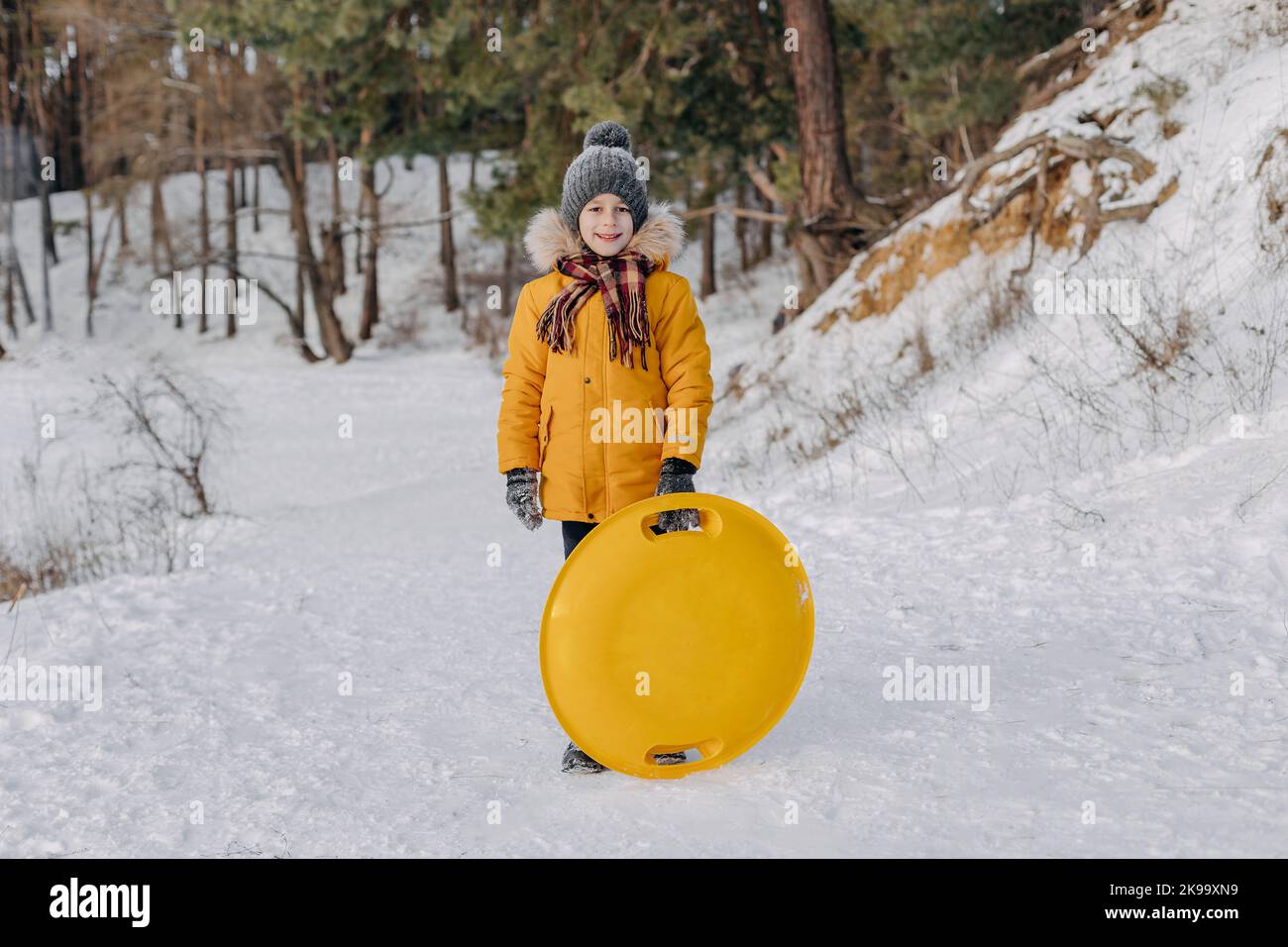 Happy fun kid holding a plastic sled on the background of snowy forest ...