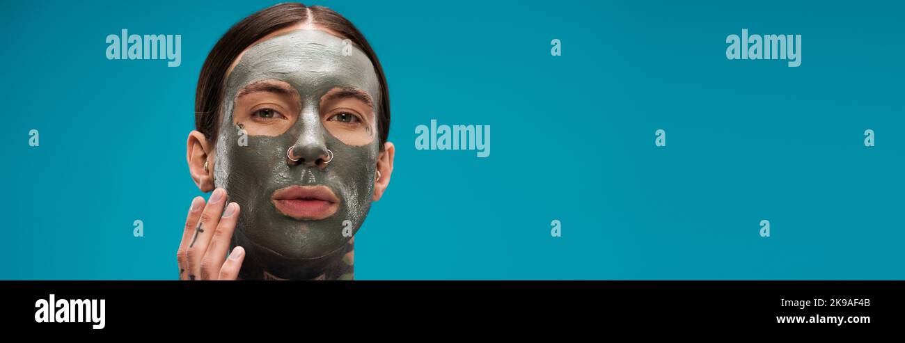 young man with tattoos touching face with clay mask isolated on blue ...