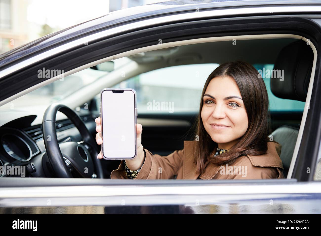 Attractive woman driving a car, she shows blank phone screen and smiles ...