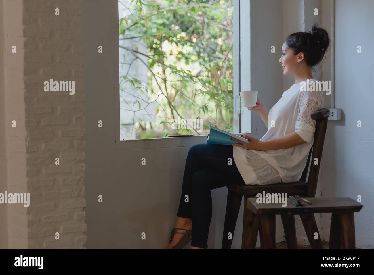 Beautiful young Asian woman drinking coffee and looking through window ...