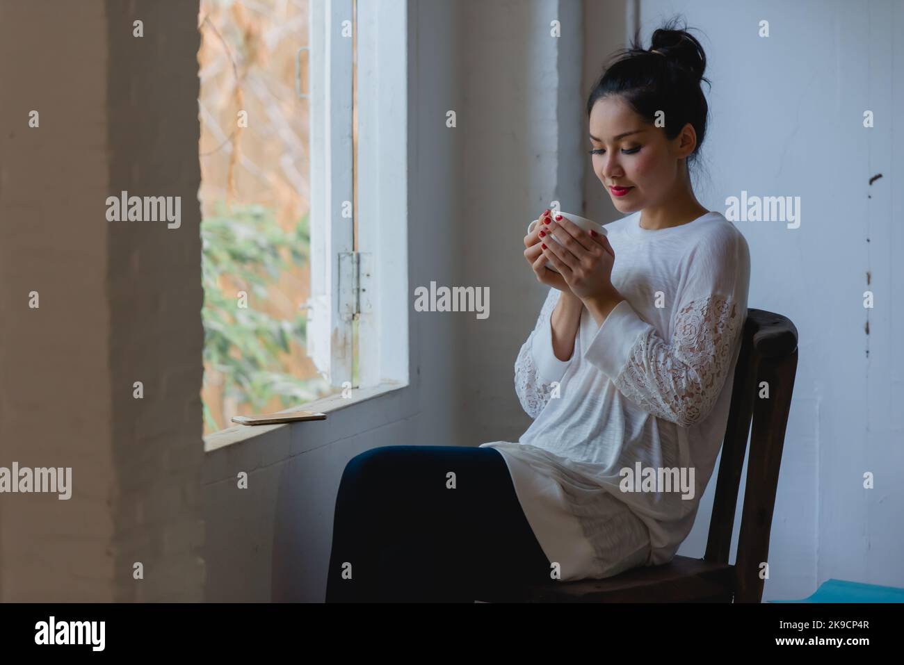 Beautiful young Asian woman drinking coffee and looking through window ...