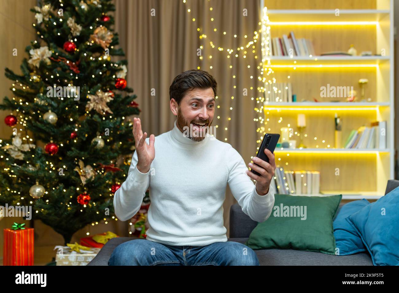 A man talks on a video call at home with a phone, sits on a sofa near a ...