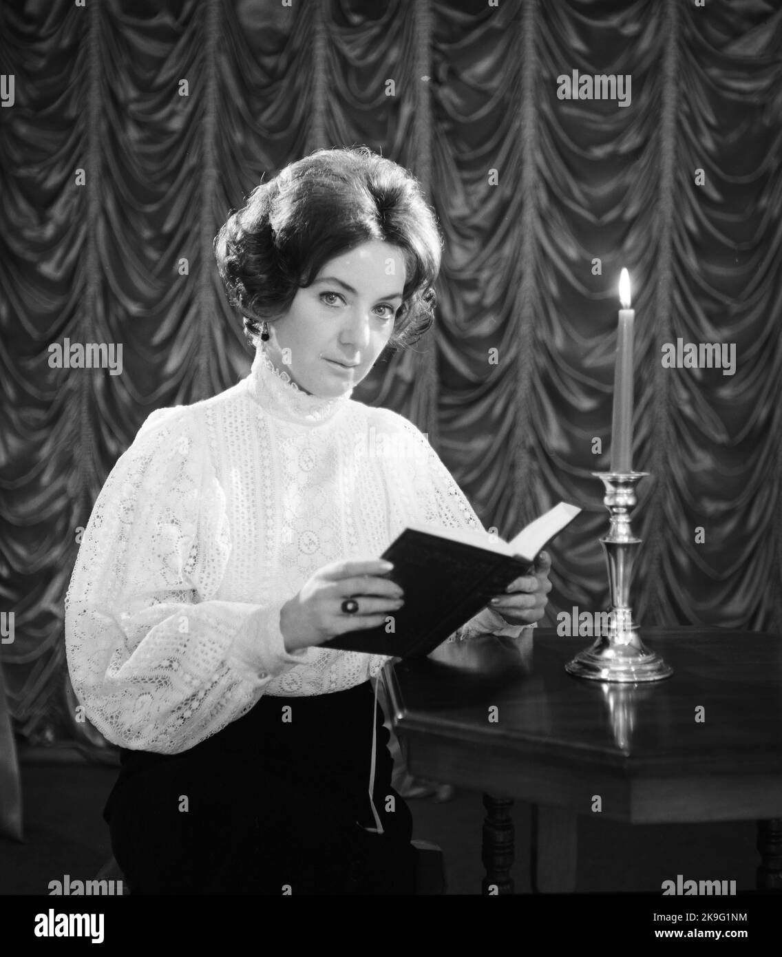 A woman in late 19th century clothing reading a book by candlelight by ...