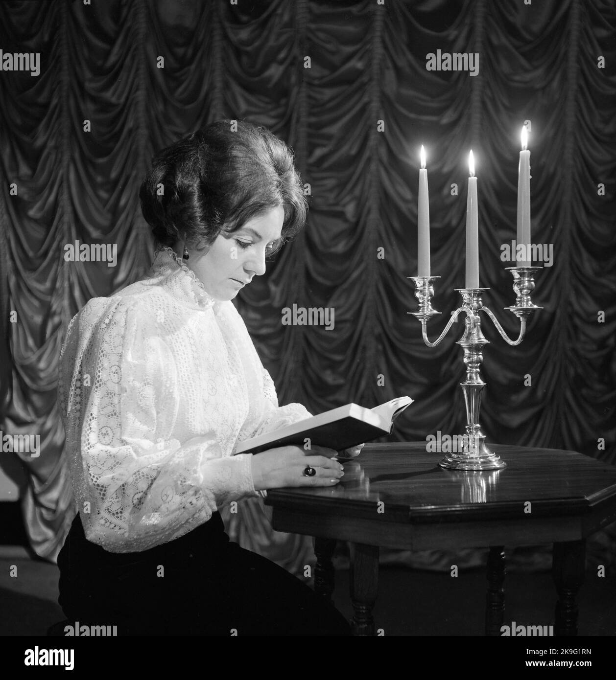 A woman in 19th century clothing reading a book by candlelight by ...