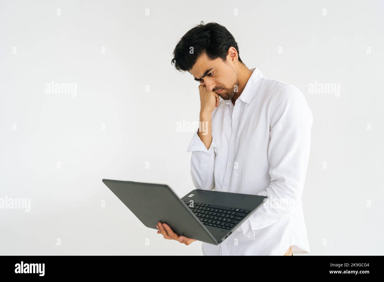 Studio shot of thoughtful young man standing with laptop looking on ...