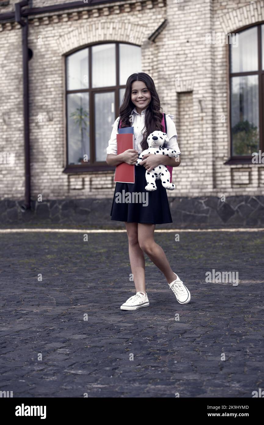 Happy small kid in school uniform hold books and toy dog in schoolyard ...