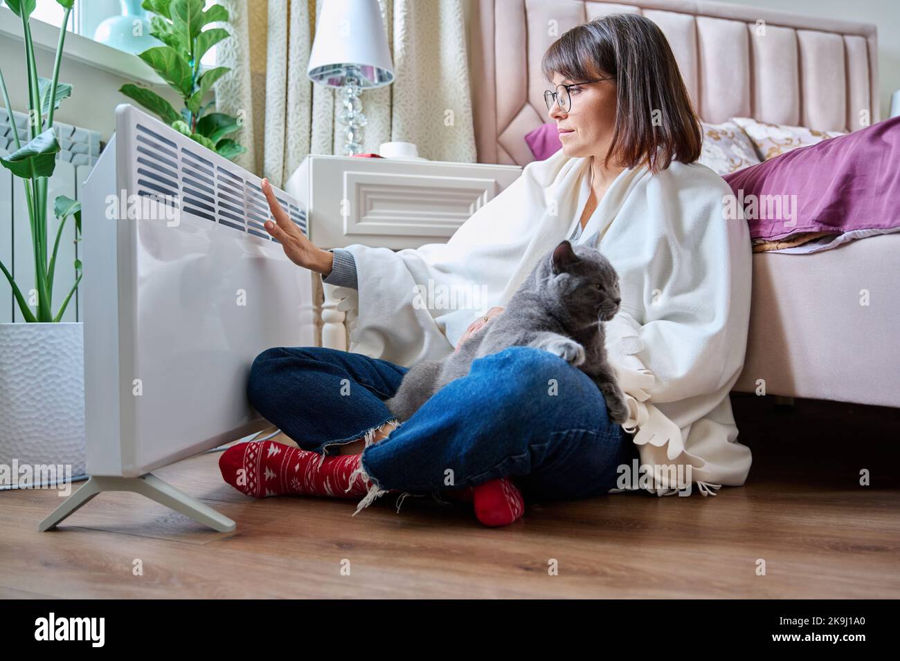 Woman sitting with cat under blanket warming near an electric heater ...