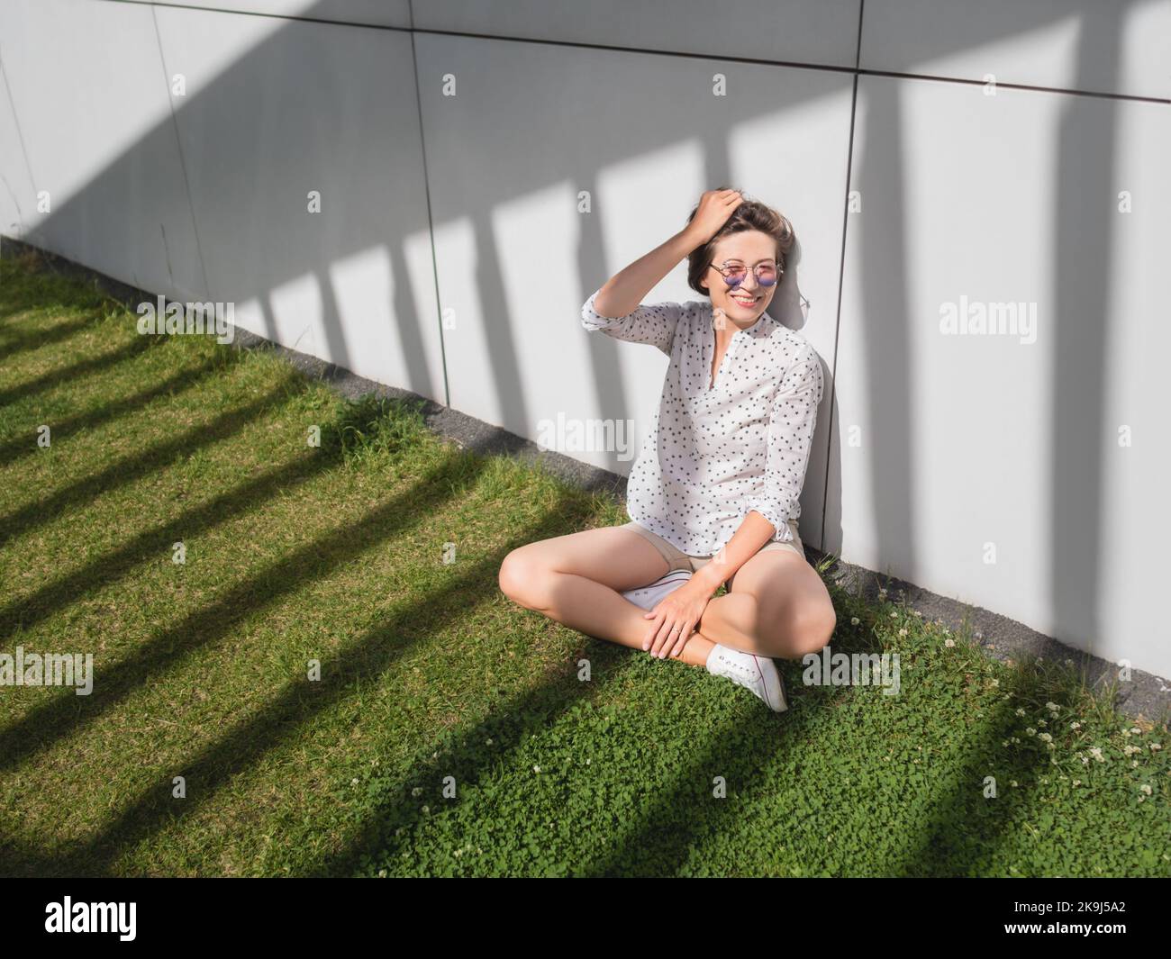 Smiling woman in colorful sunglasses has a rest on lawn in urban park. Nature in town. Relax ...