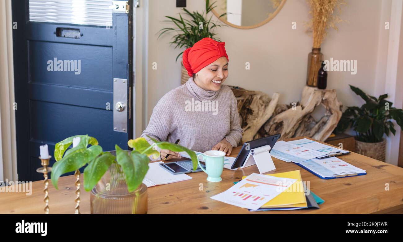 Happy biracial woman wearing hijab, sitting at table in living room ...