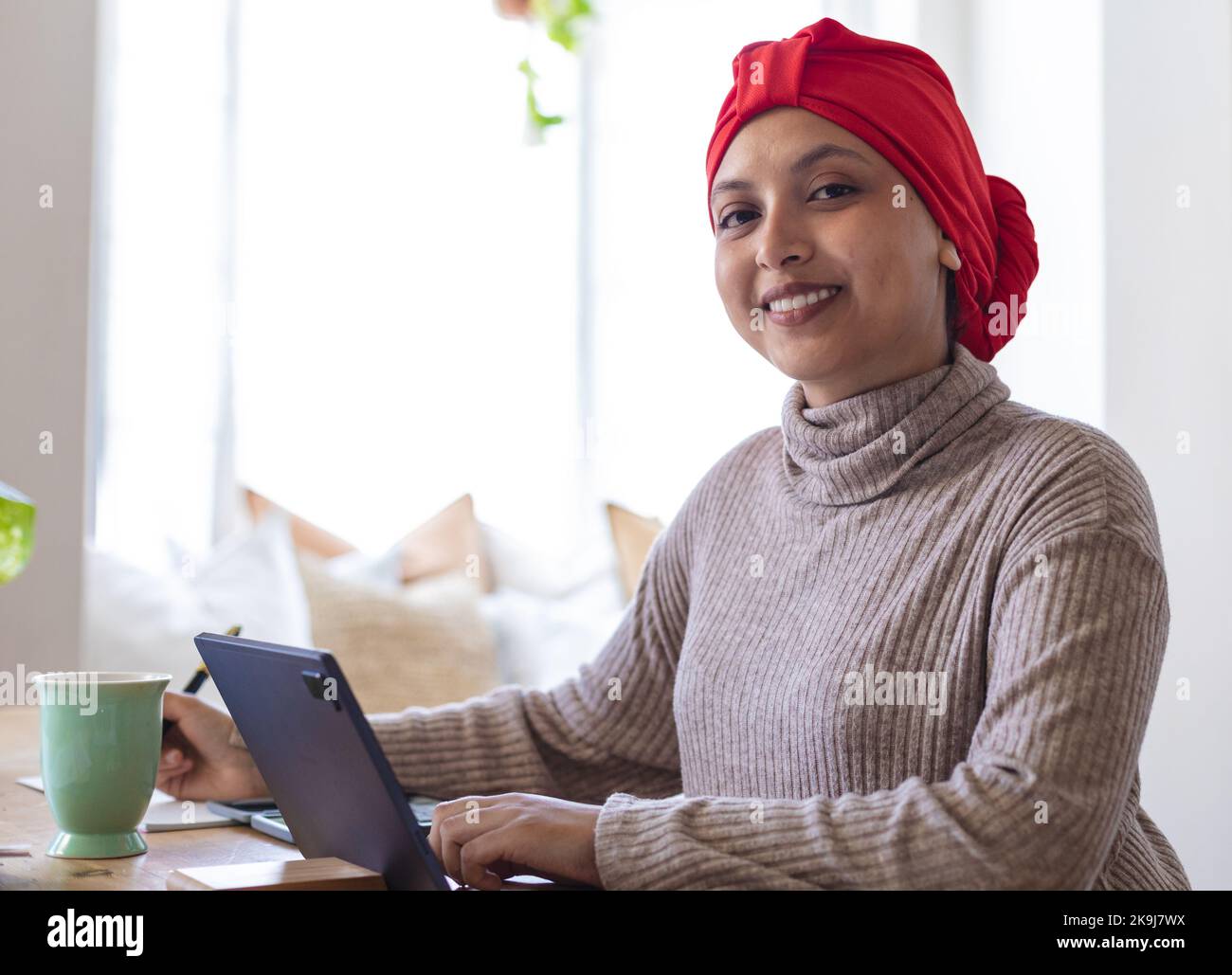 Portrait of happy biracial woman wearing hijab, sitting at table, using ...