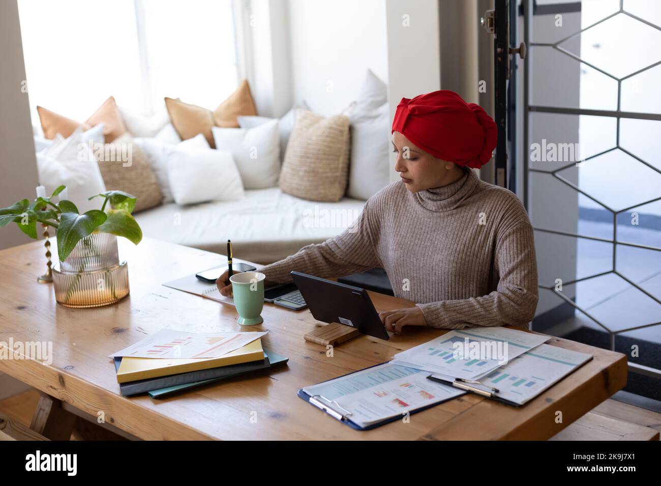 Happy biracial woman wearing hijab, sitting at table in living room ...