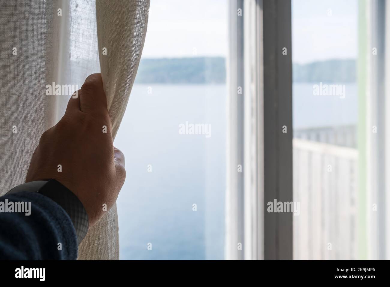 Hand draws back the curtain on the window, through which a view of the ...