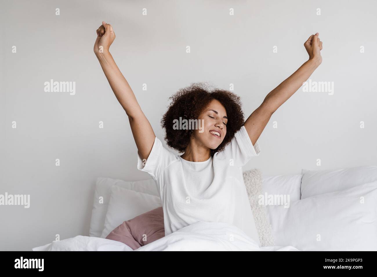 African girl in pajamas stertching in cozy bed and raising hands up at ...