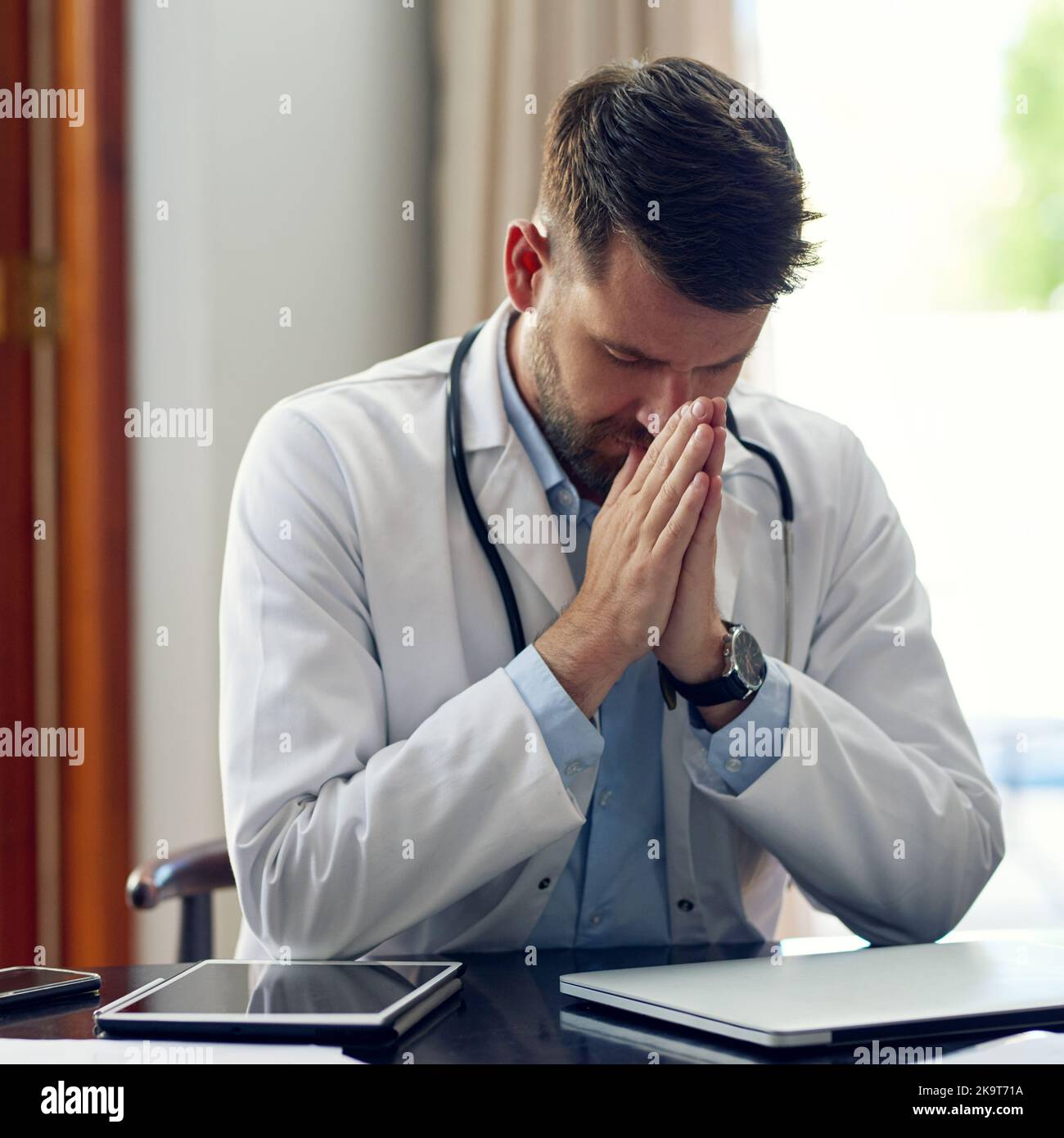 Praying for a miracle. a handsome male doctor praying while sitting in ...