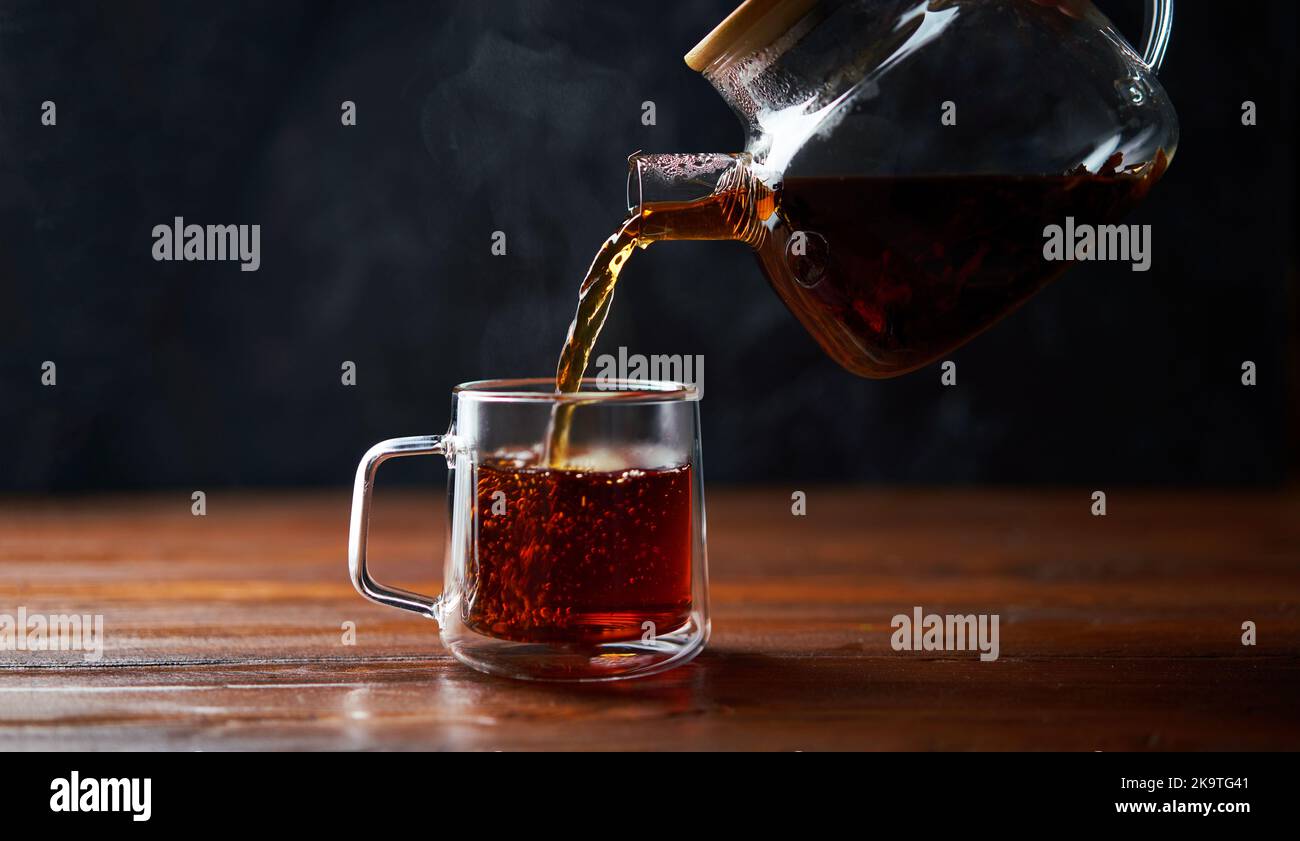 Tea poured from a teapot into a cup standing on a brown wooden table ...