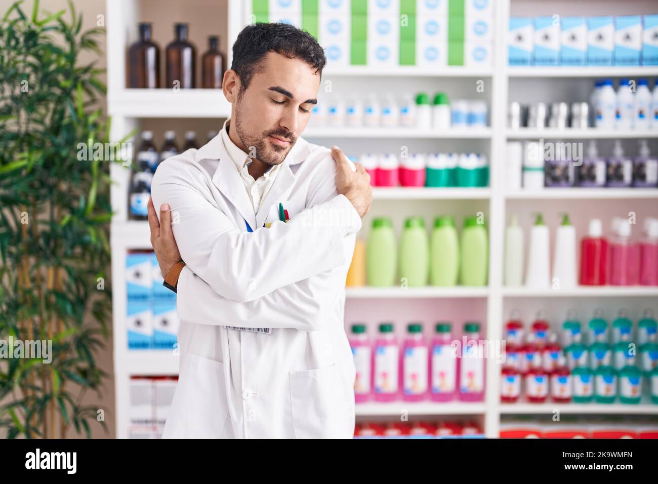 Handsome hispanic man working at pharmacy drugstore hugging oneself ...