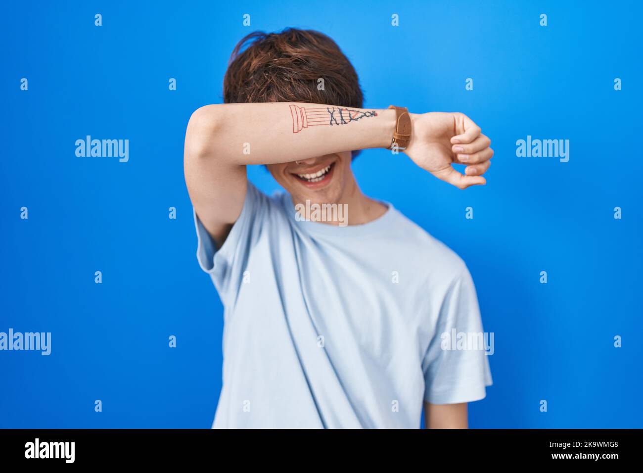 Hispanic young man standing over blue background covering eyes with arm ...