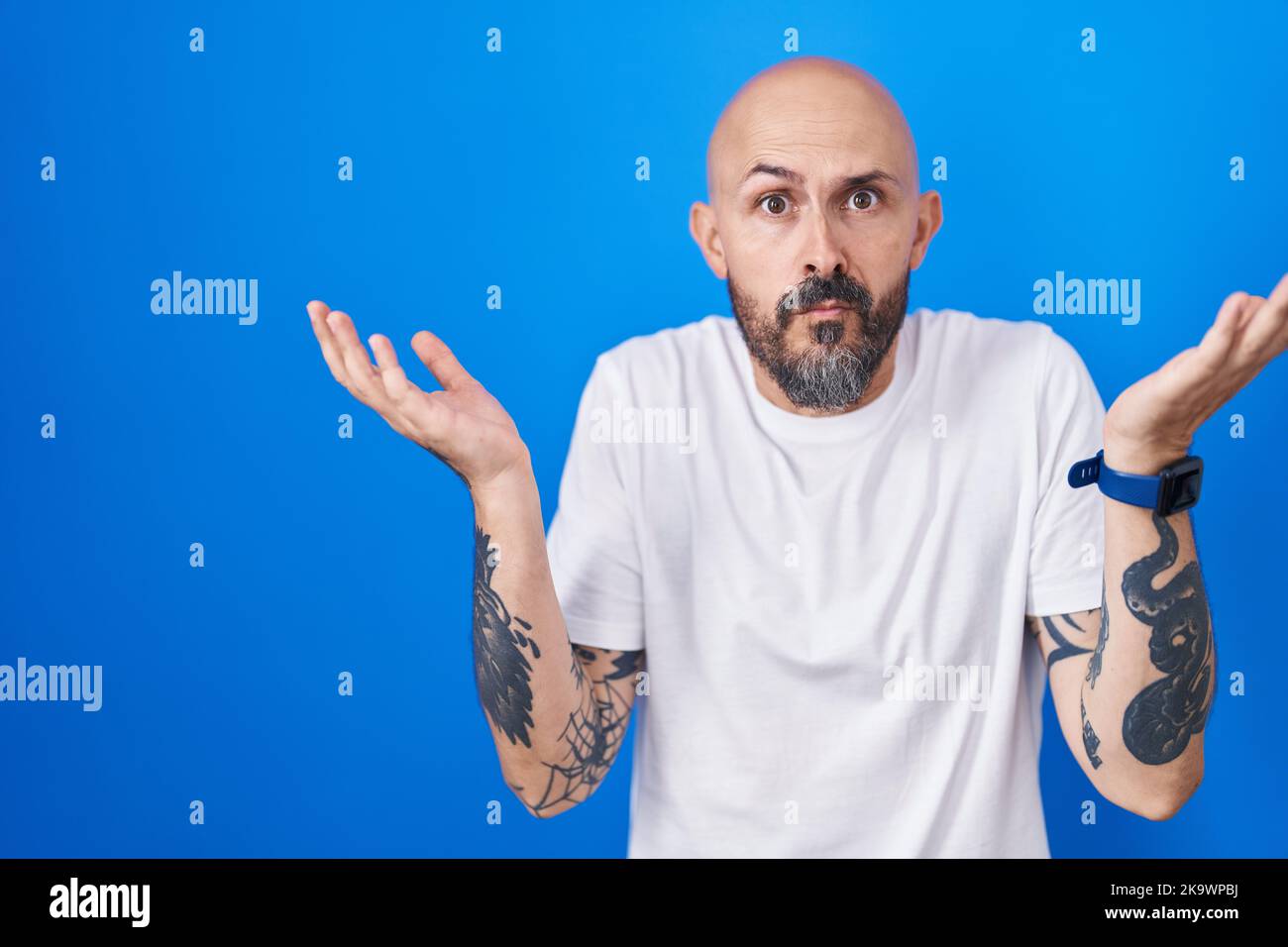 Hispanic man with tattoos standing over blue background clueless and ...