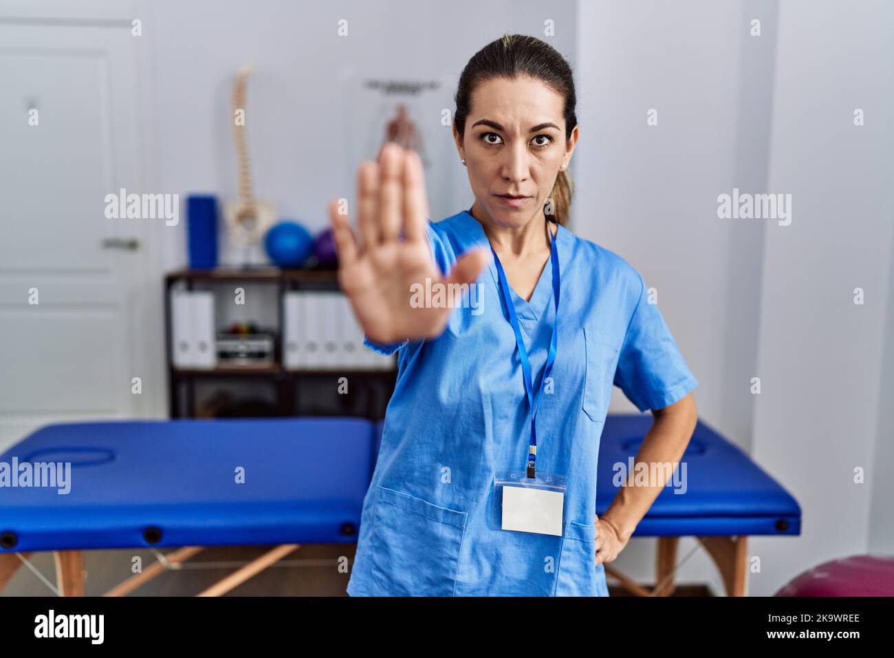 Young hispanic woman wearing physiotherapist uniform standing at clinic ...