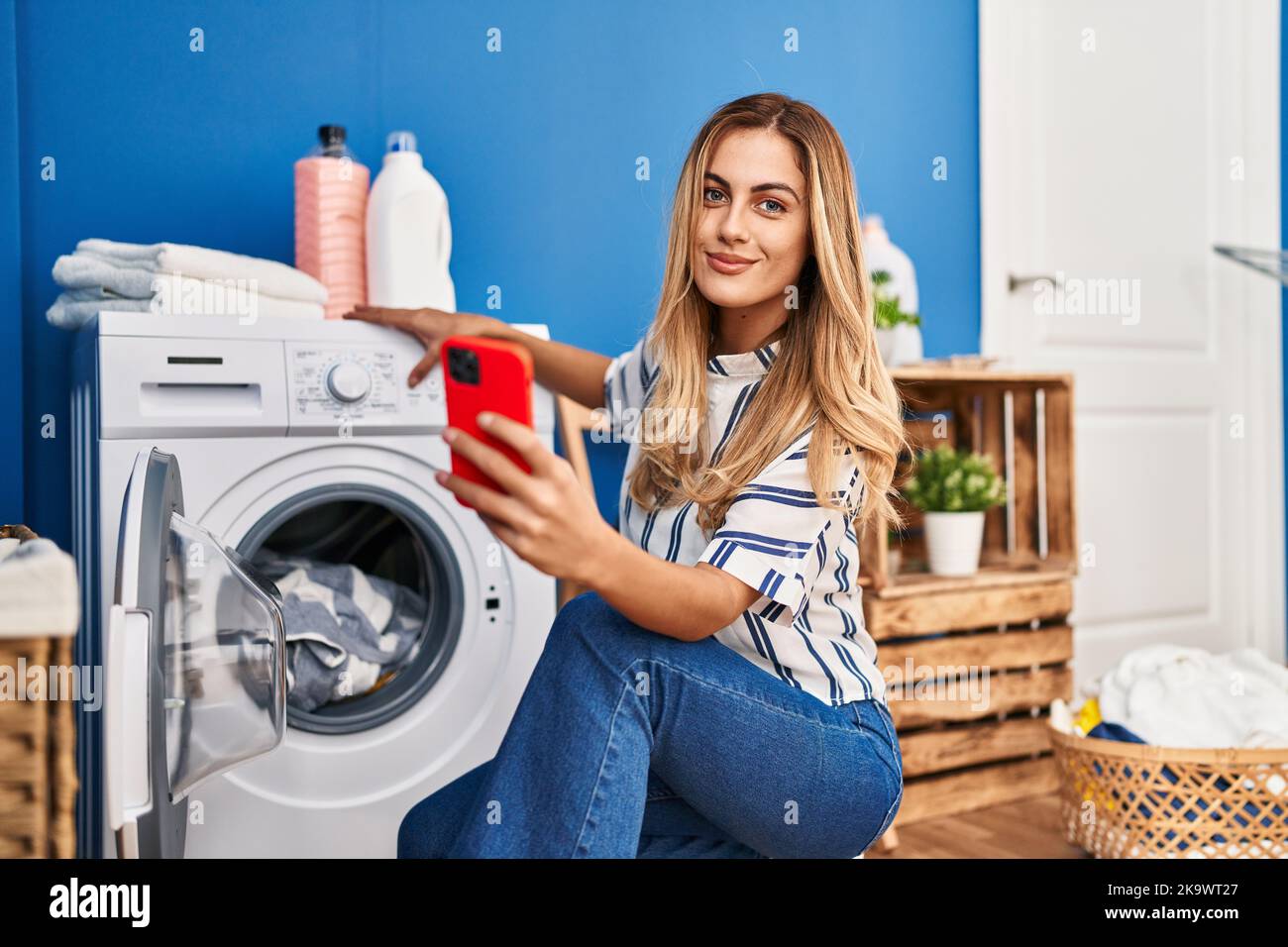 Young blonde woman using smartphone and washing clothes at laundry room ...