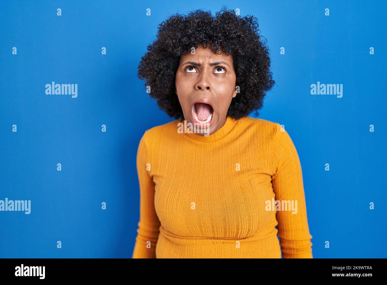 Black woman with curly hair standing over blue background angry and mad ...