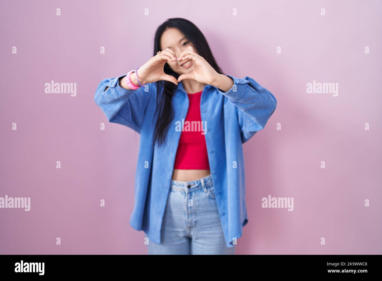 Young asian woman standing over pink background smiling in love doing ...