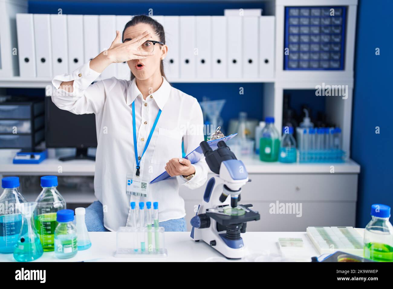 Young brunette woman working at scientist laboratory peeking in shock ...