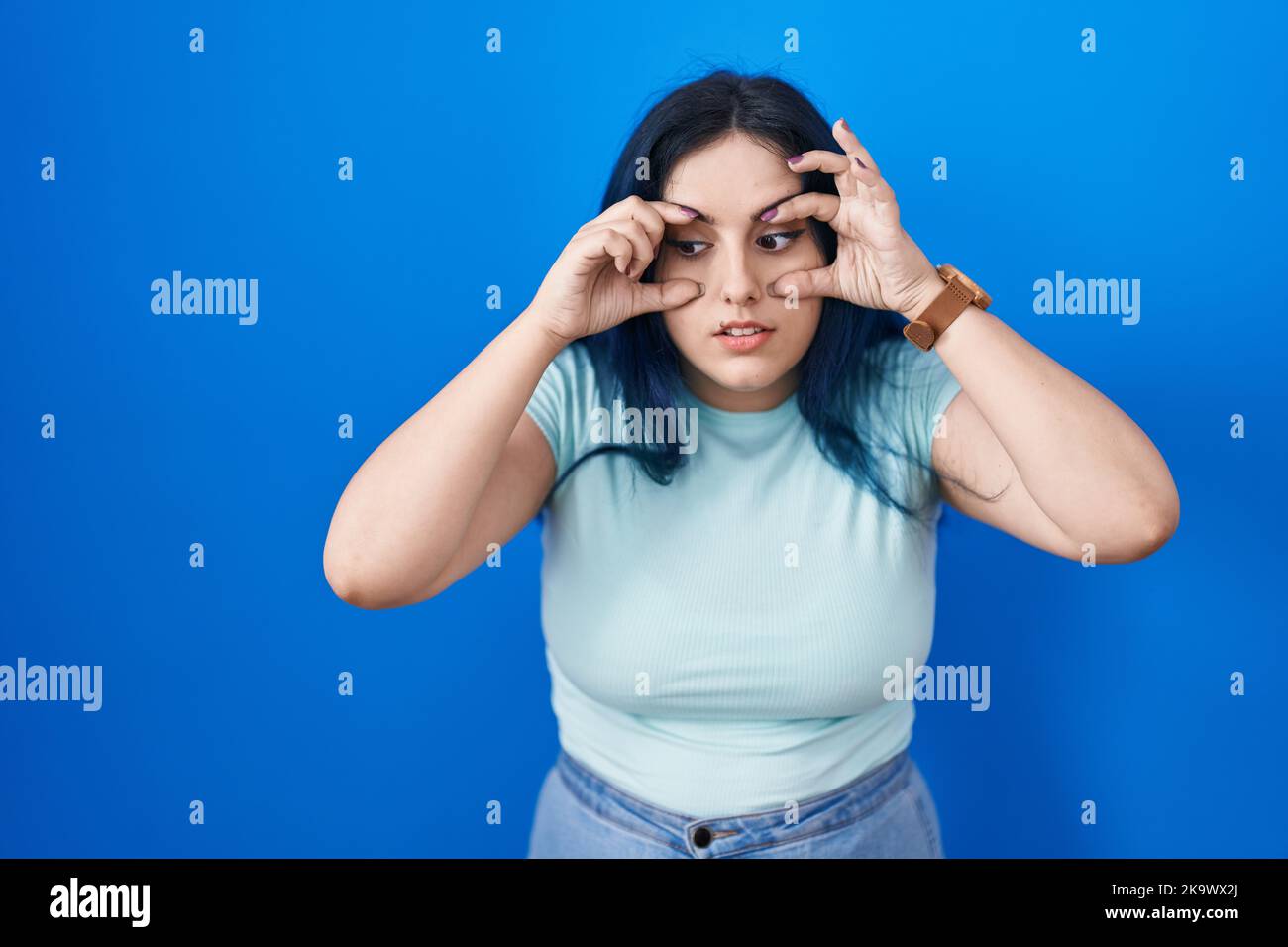 Young modern girl with blue hair standing over blue background trying ...