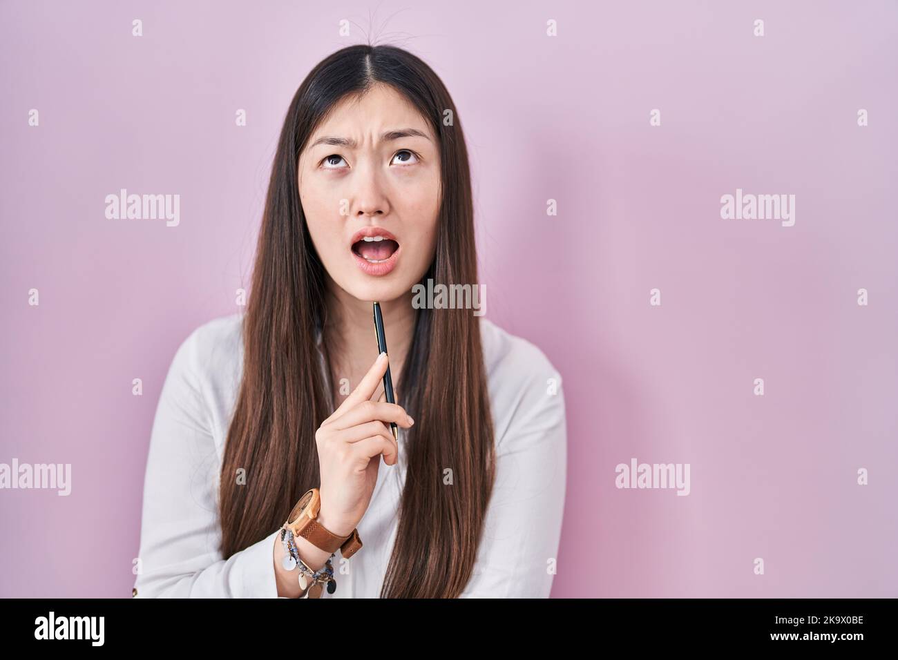 Chinese young woman holding pencil over pink background angry and mad screaming frustrated and ...