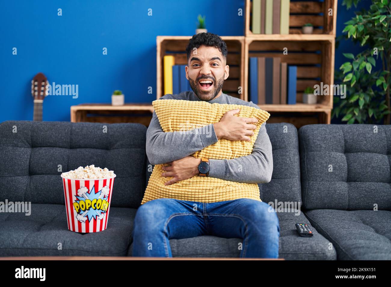 Hispanic man with beard eating popcorn watching a movie at home smiling ...