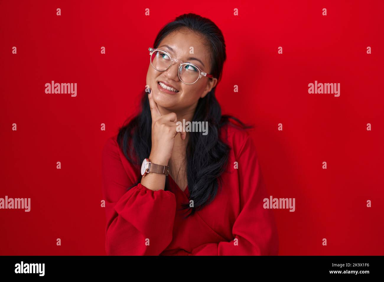 Asian young woman standing over red background with hand on chin ...