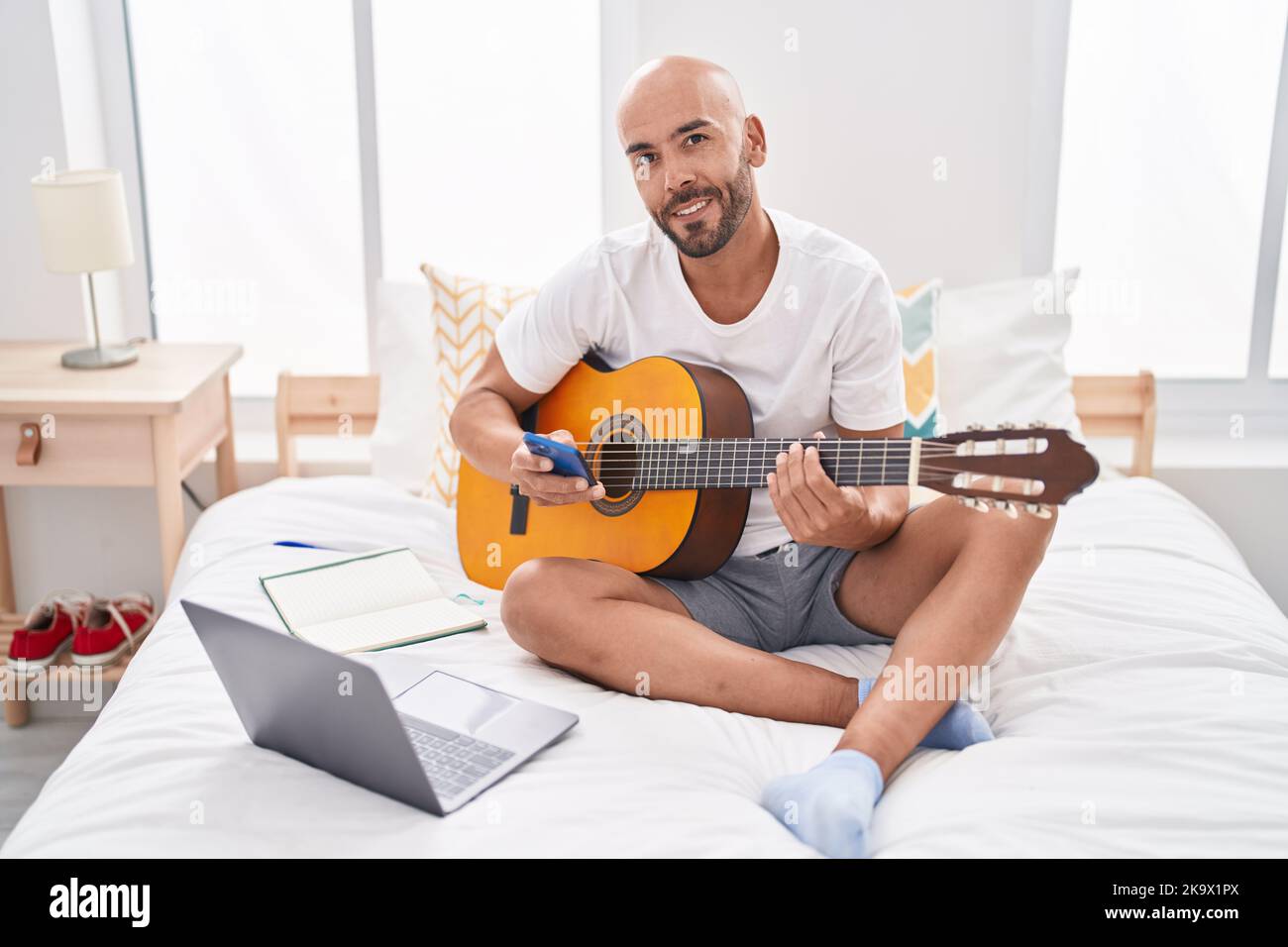 Young bald man playing classical guitar using smartphone at bedroom ...