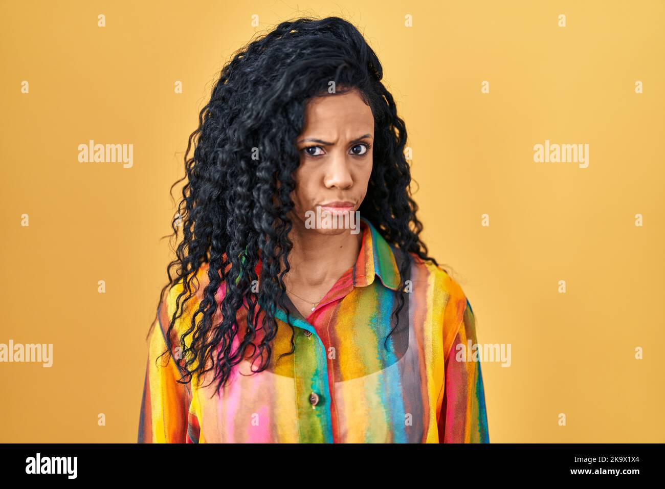 Middle age hispanic woman standing over yellow background skeptic and ...