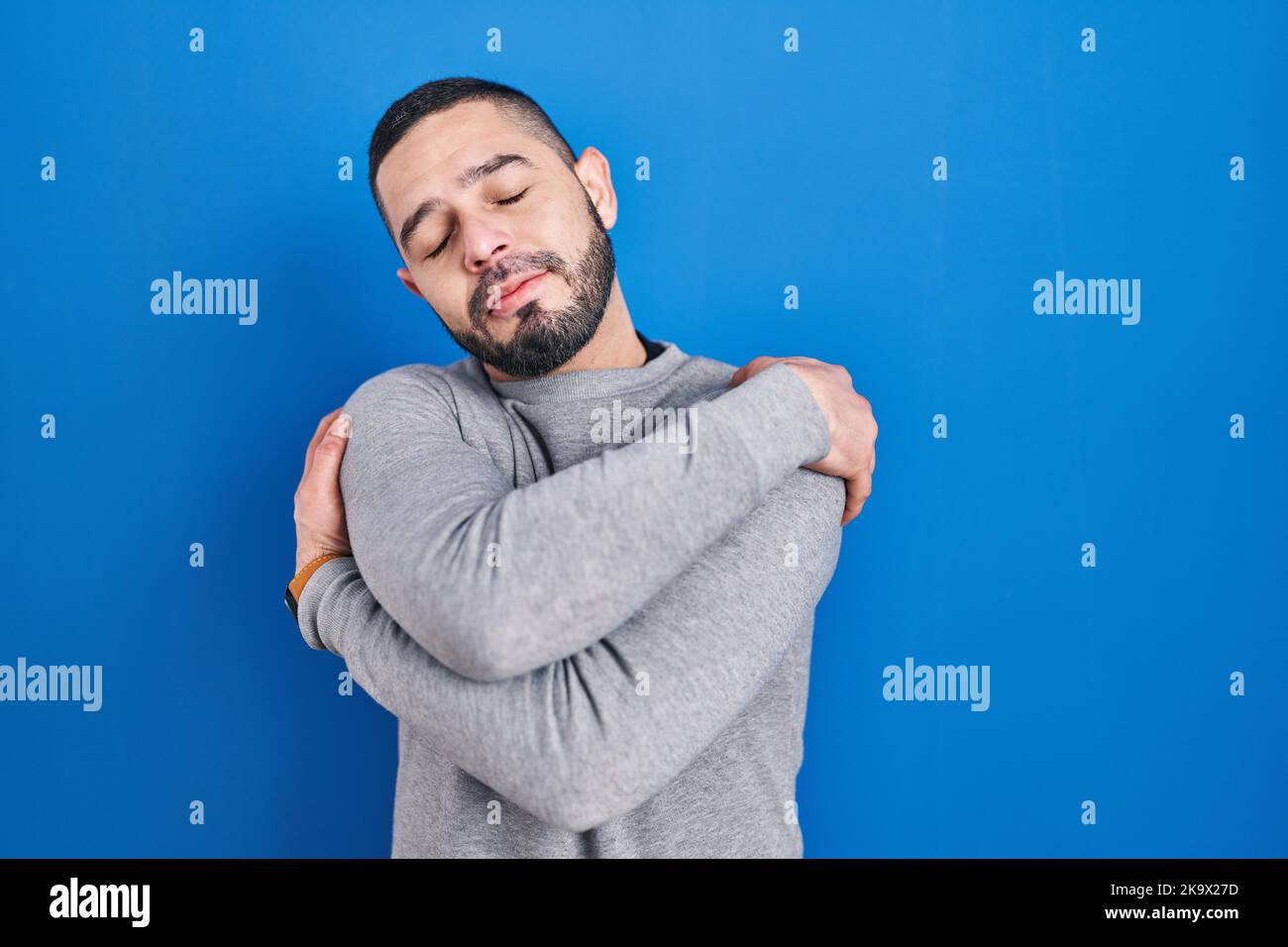 Hispanic man standing over blue background hugging oneself happy and ...