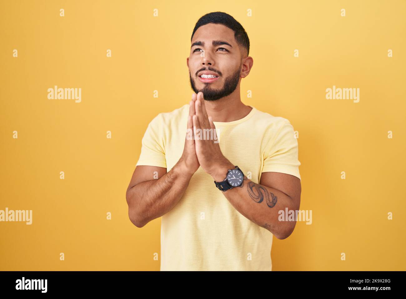 Young hispanic man standing over yellow background begging and praying ...