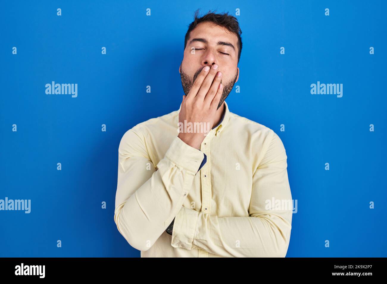 Handsome hispanic man standing over blue background bored yawning tired ...