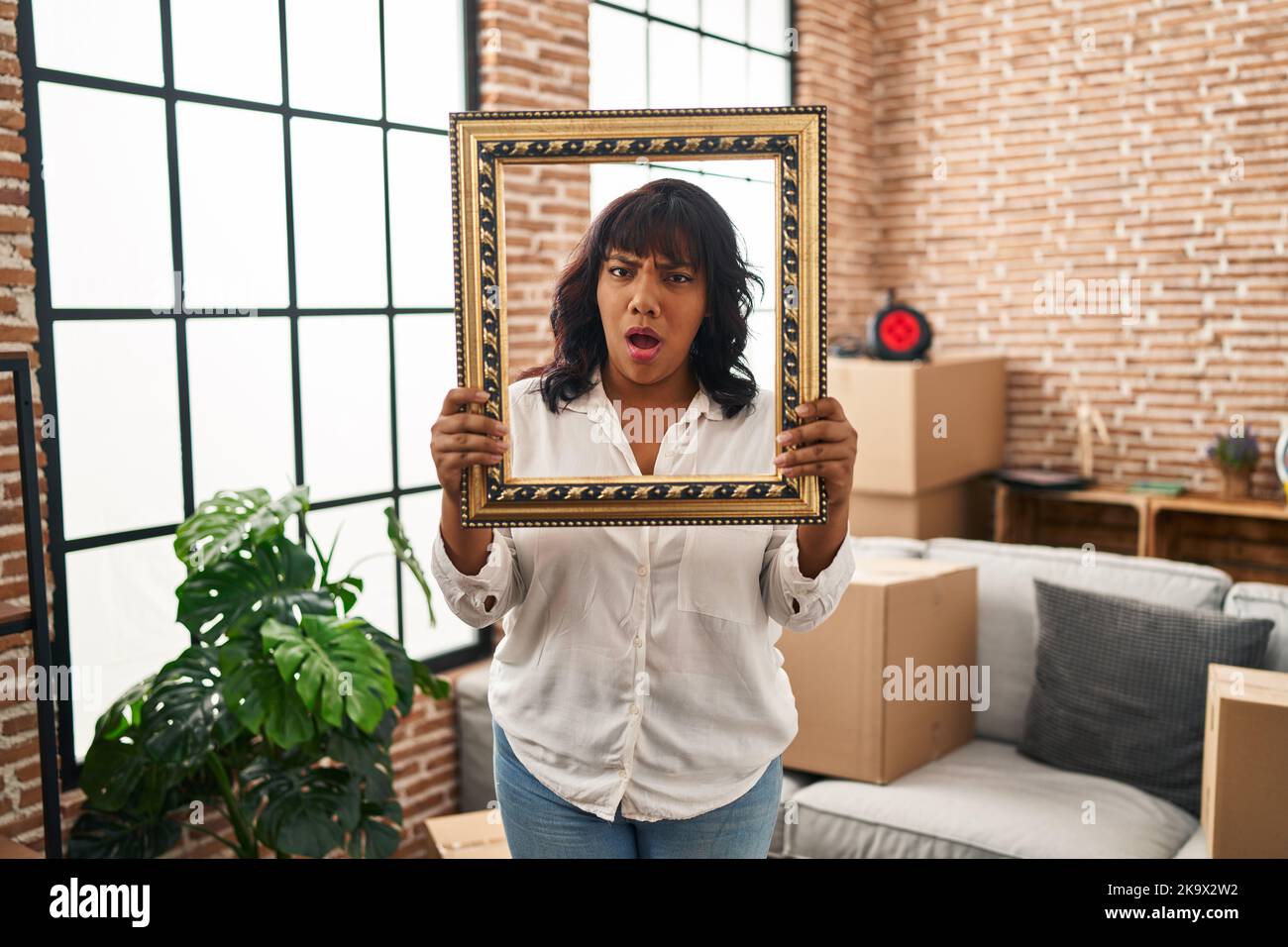 Hispanic woman at new home holding empty frame in shock face, looking ...