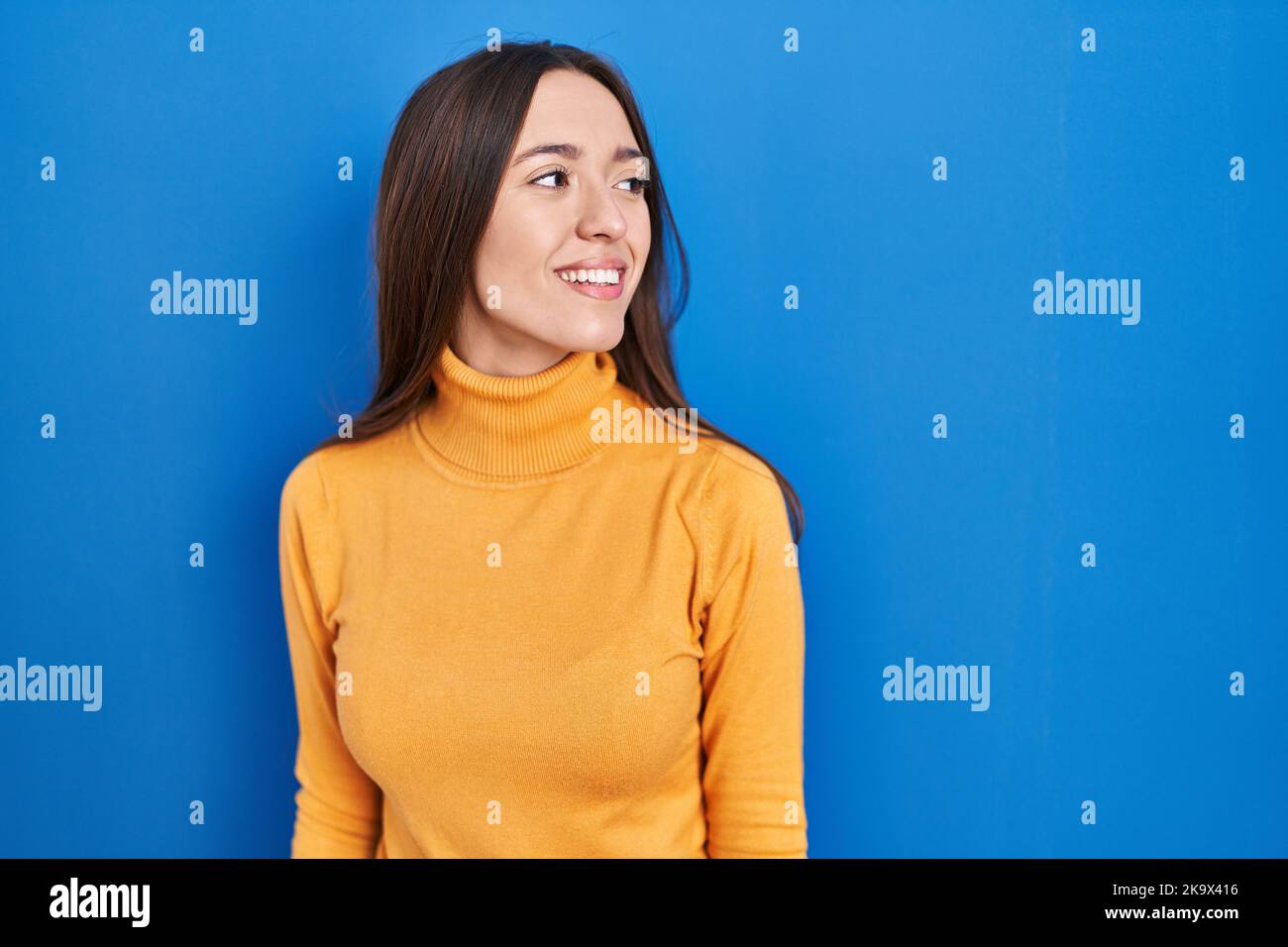 Young brunette woman standing over blue background looking away to side ...